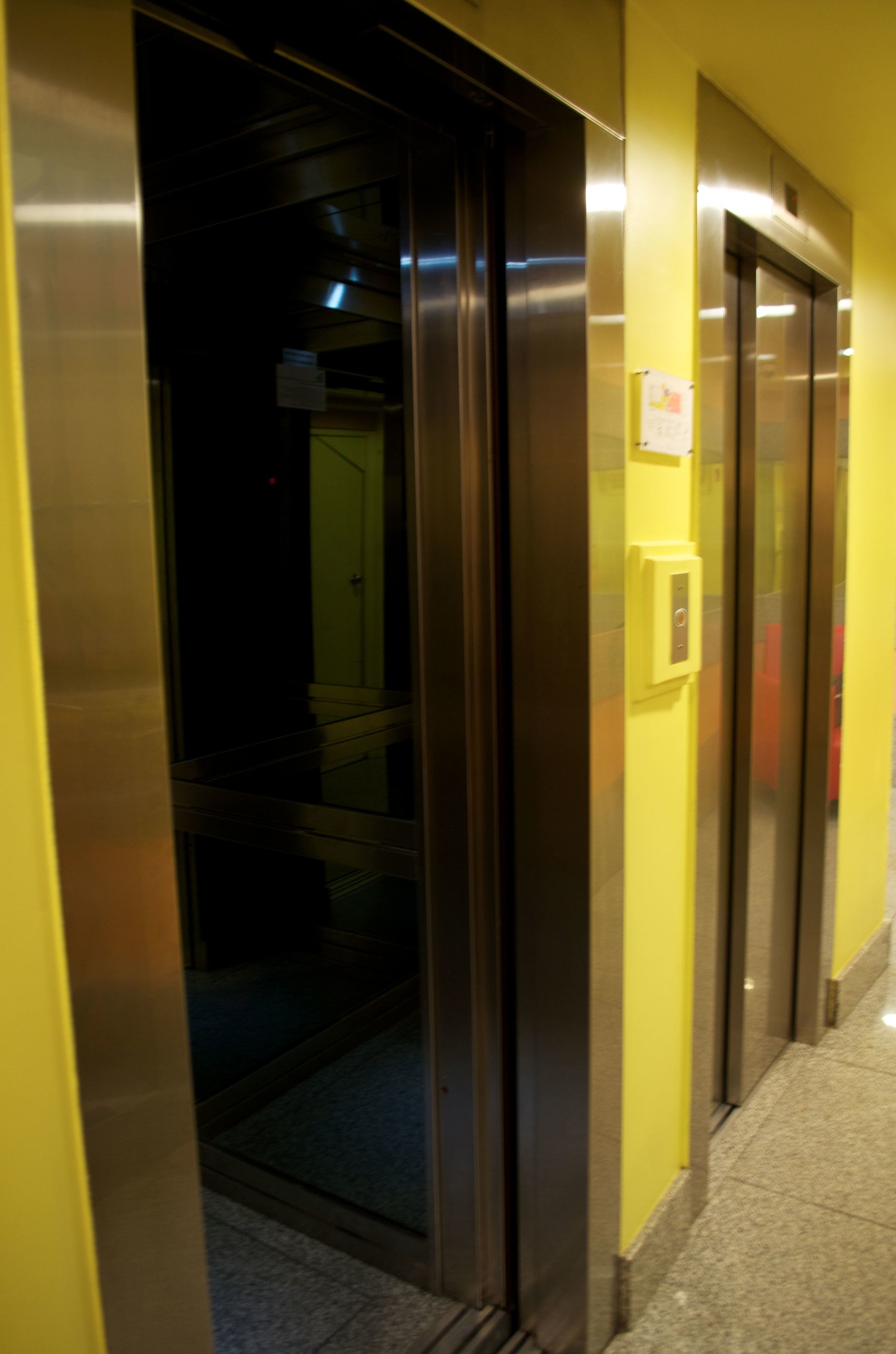 Um elevador em um corredor com paredes amarelas e uma porta metálica.