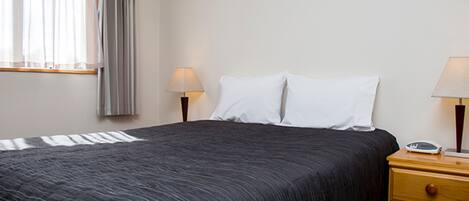 Quarto de hotel com uma cama grande, duas mesas de cabeceira com luminárias, uma janela com cortinas e uma pequena mesinha de madeira.