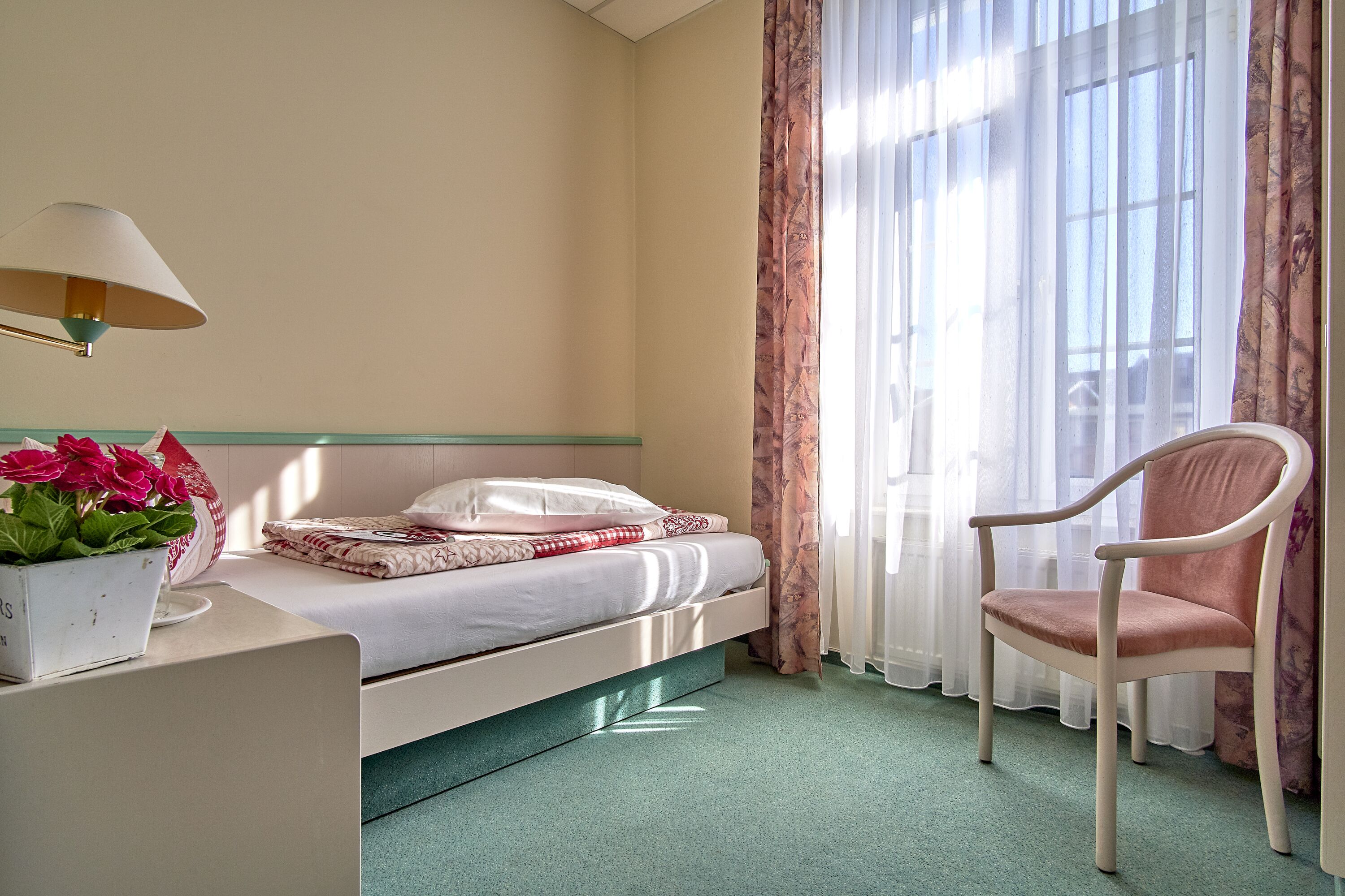 Quarto de hotel com cama, uma cadeira, escrivaninha com luminária e janela com cortinas.