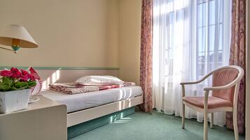 Quarto de hotel com cama, uma cadeira, escrivaninha com luminária e janela com cortinas.