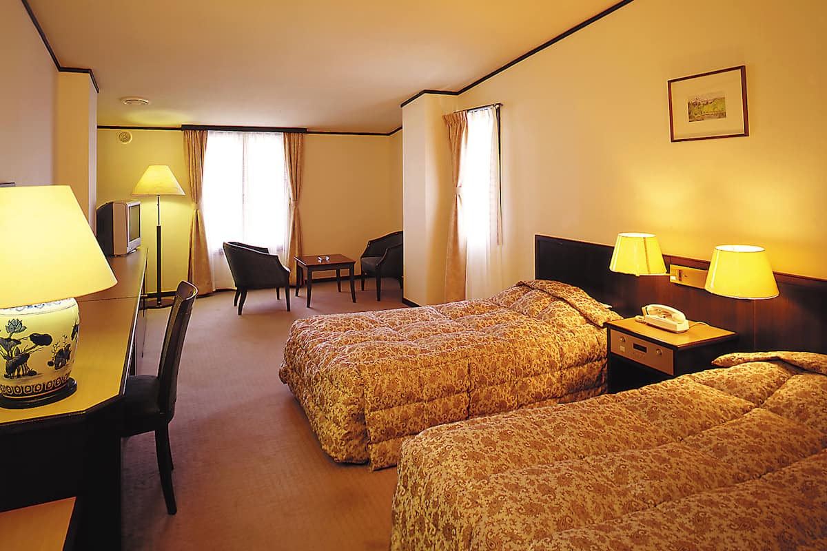 Quarto de hotel com duas camas, uma escrivaninha com luminária, uma cadeira, uma mesinha e uma televisão.