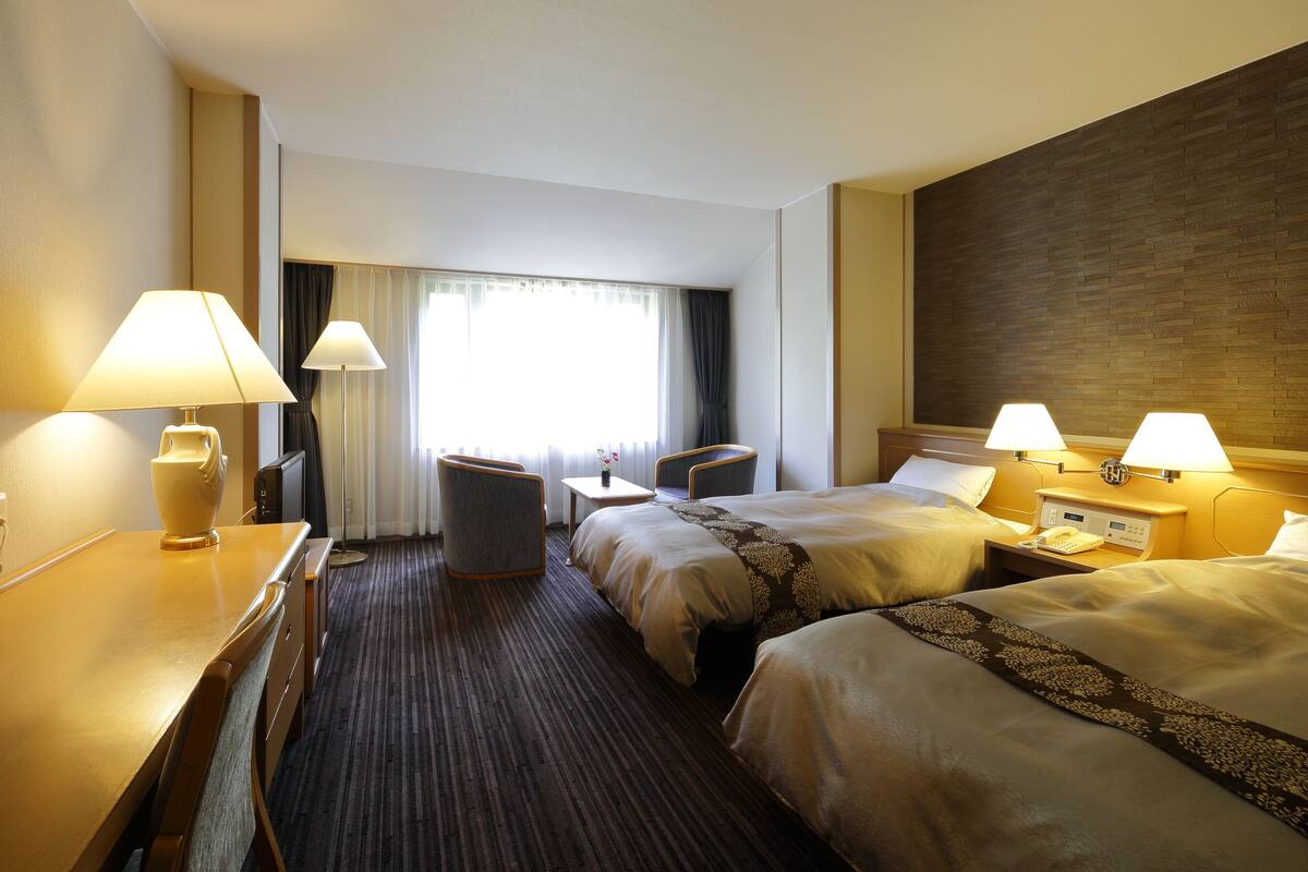 Quarto de hotel com duas camas, uma escrivaninha com luminária, uma cadeira, uma mesinha de cabeceira com luminária e uma janela com cortinas.