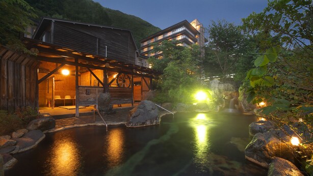 Um ryokan japonês tradicional com fachada de madeira, banho termal de pedra e vista para uma montanha ao fundo.