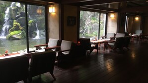 Quarto com piso de madeira, grandes janelas com vista para uma cascata, e várias cadeiras e mesas.