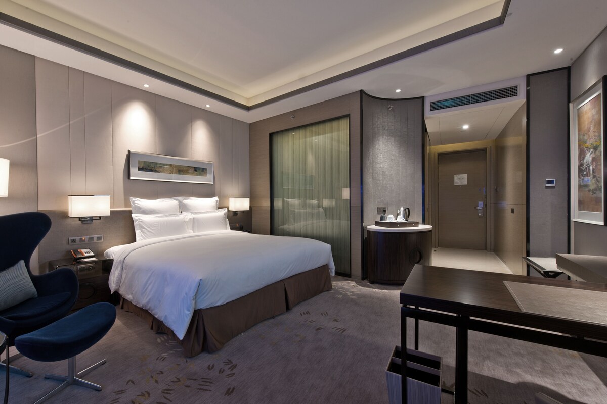 Quarto de hotel moderno com uma cama grande, uma escrivaninha, uma cadeira e uma mesa de cabeceira.