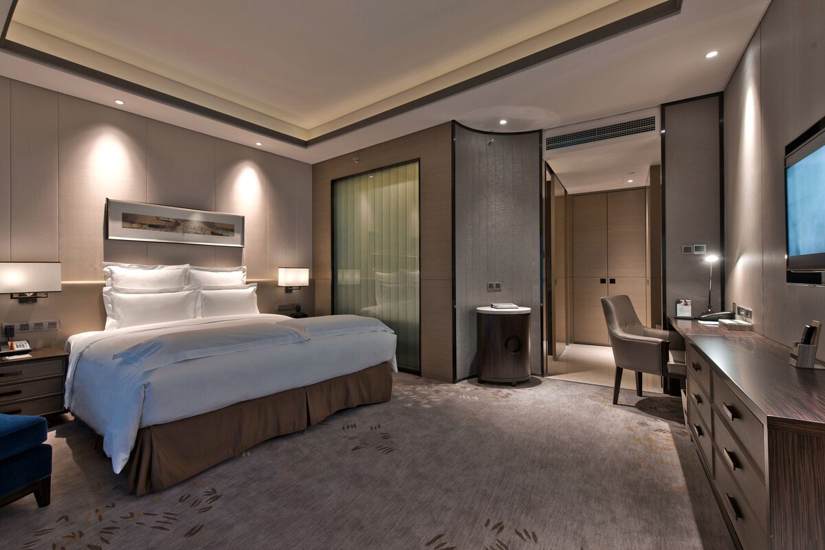 Quarto de hotel moderno com uma cama grande, mesas de cabeceira, uma escrivaninha, uma cadeira, uma televisão e uma janela ampla.