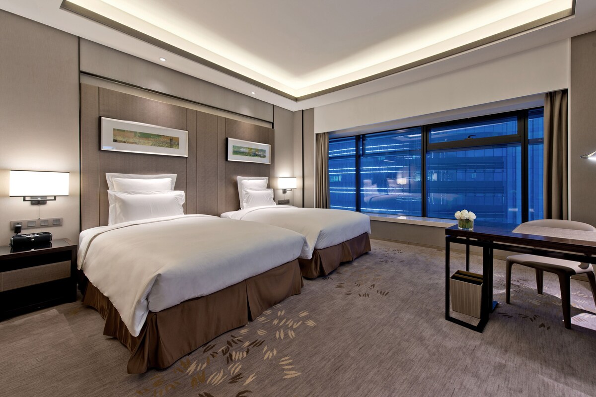 Quarto de hotel com duas camas, uma escrivaninha, uma cadeira, uma janela com vista para a cidade e uma luminária.