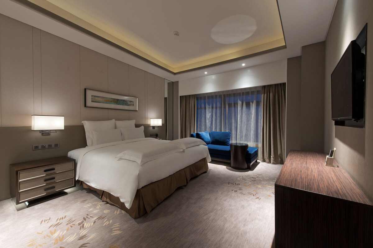 Quarto de hotel moderno com uma cama grande, escrivaninha, televisão e janela com cortinas.