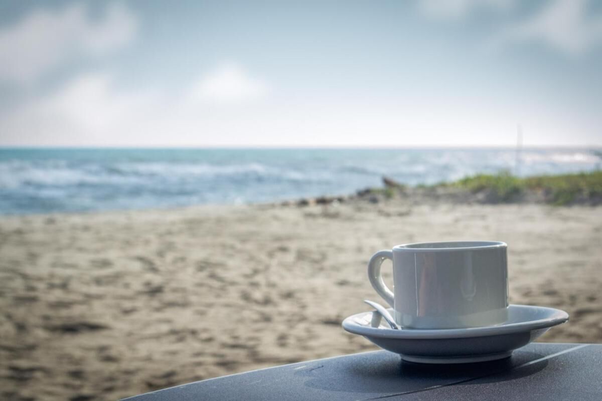 Xícara e pires brancos sobre uma mesa, com vista para o oceano e a praia ao fundo.
