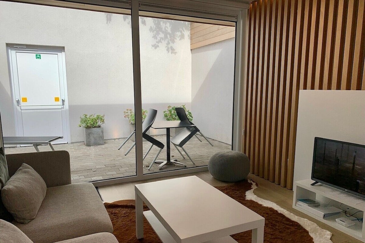 Uma sala de estar moderna com sofá, mesa de centro, televisão e porta de vidro deslizante que dá para uma área externa com cadeiras e plantas.