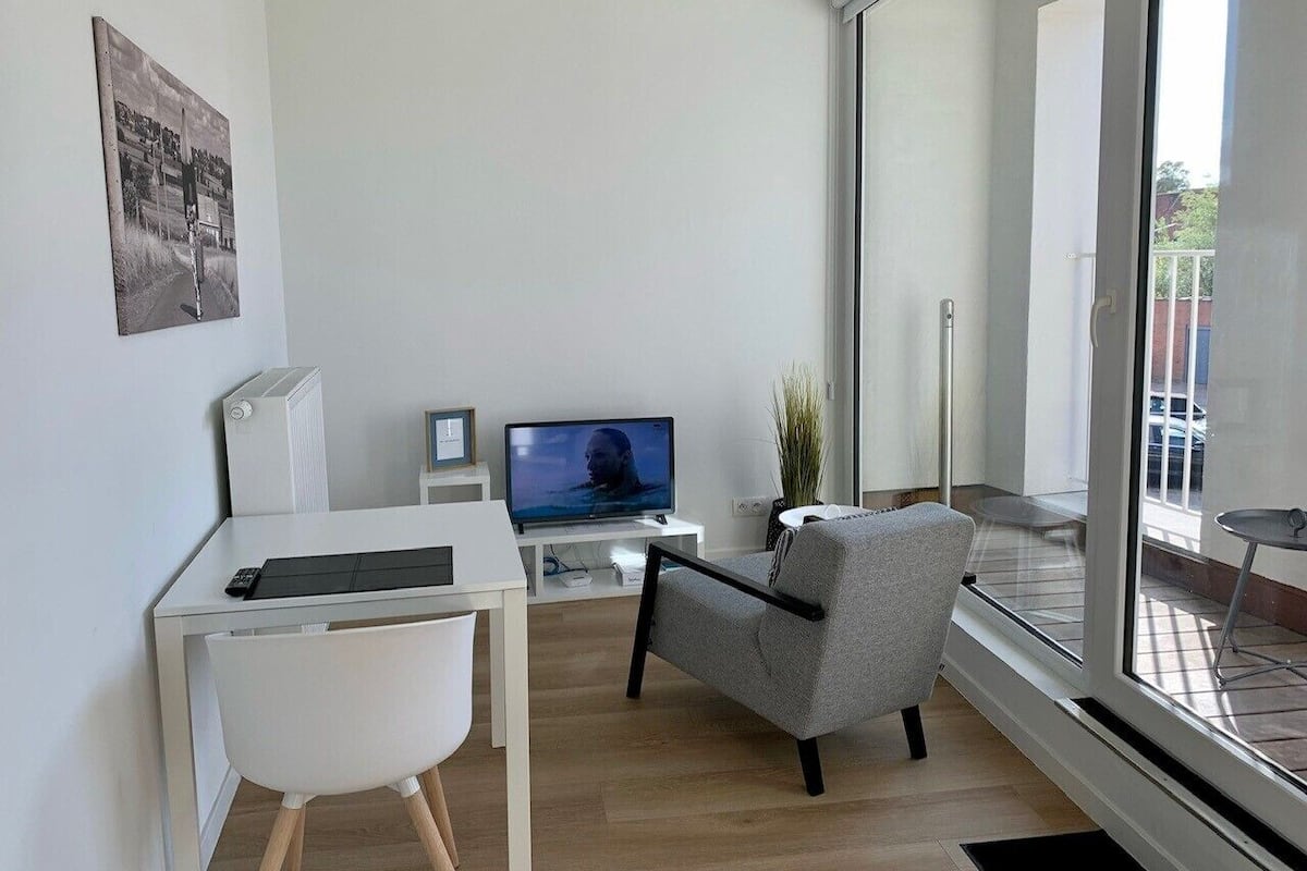 Uma sala de estar moderna com uma escrivaninha branca, uma poltrona cinza, uma televisão e uma janela ampla com vista para o exterior.