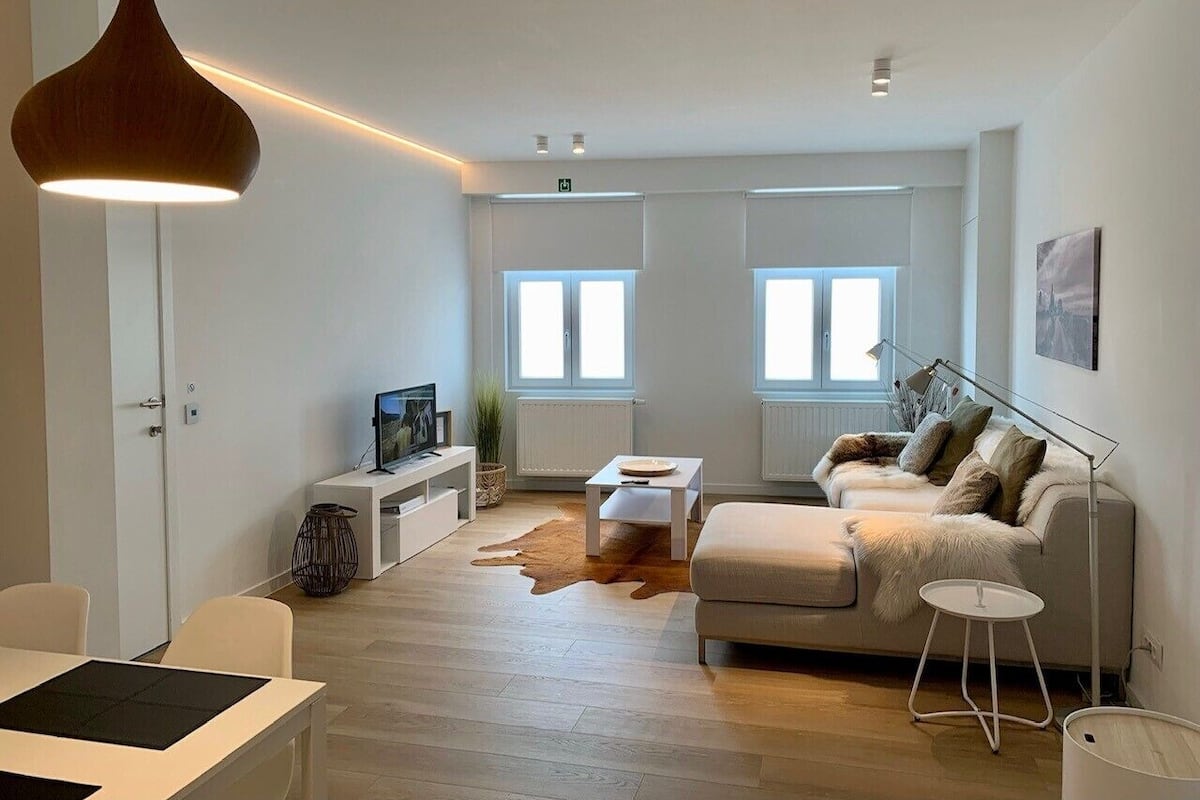 Uma sala de estar moderna com sofá, mesa de centro e televisão. Há janelas com persianas, um candeeiro pendente e um radiador.