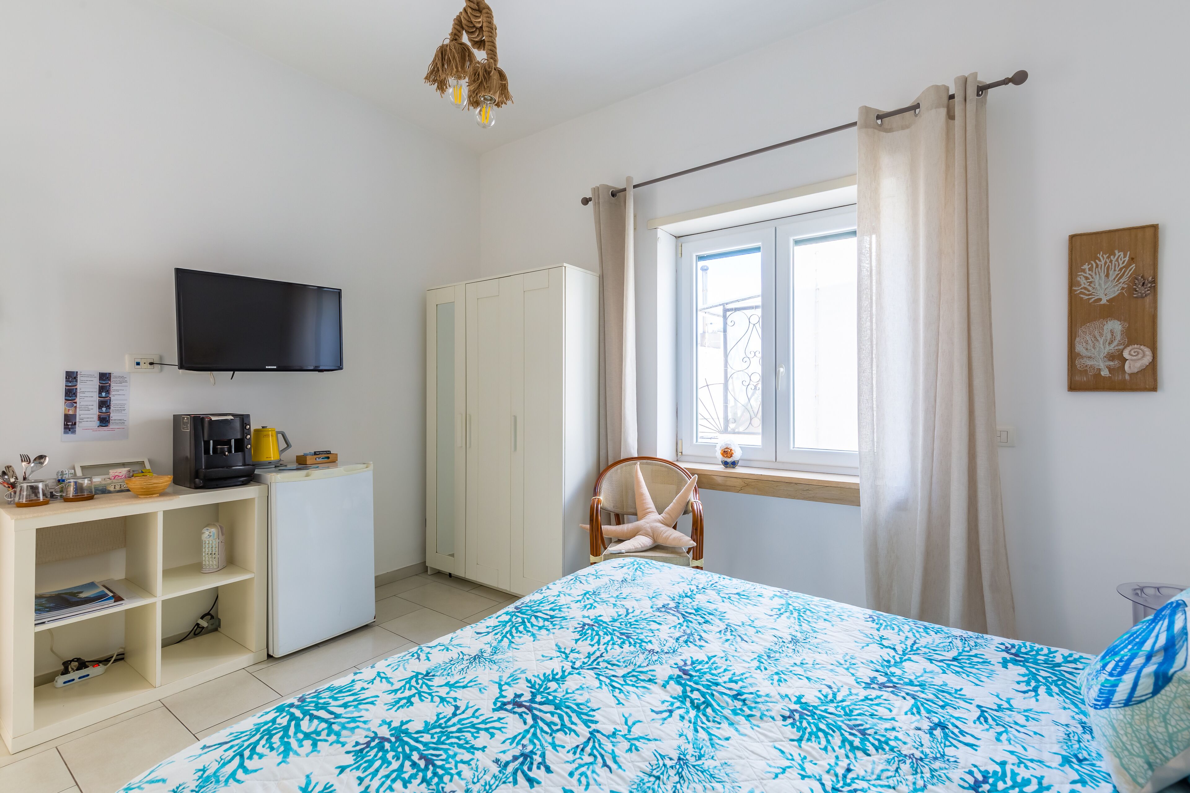Quarto com cama, televisão, secretária com máquina de café, cadeira e janela com cortinas.