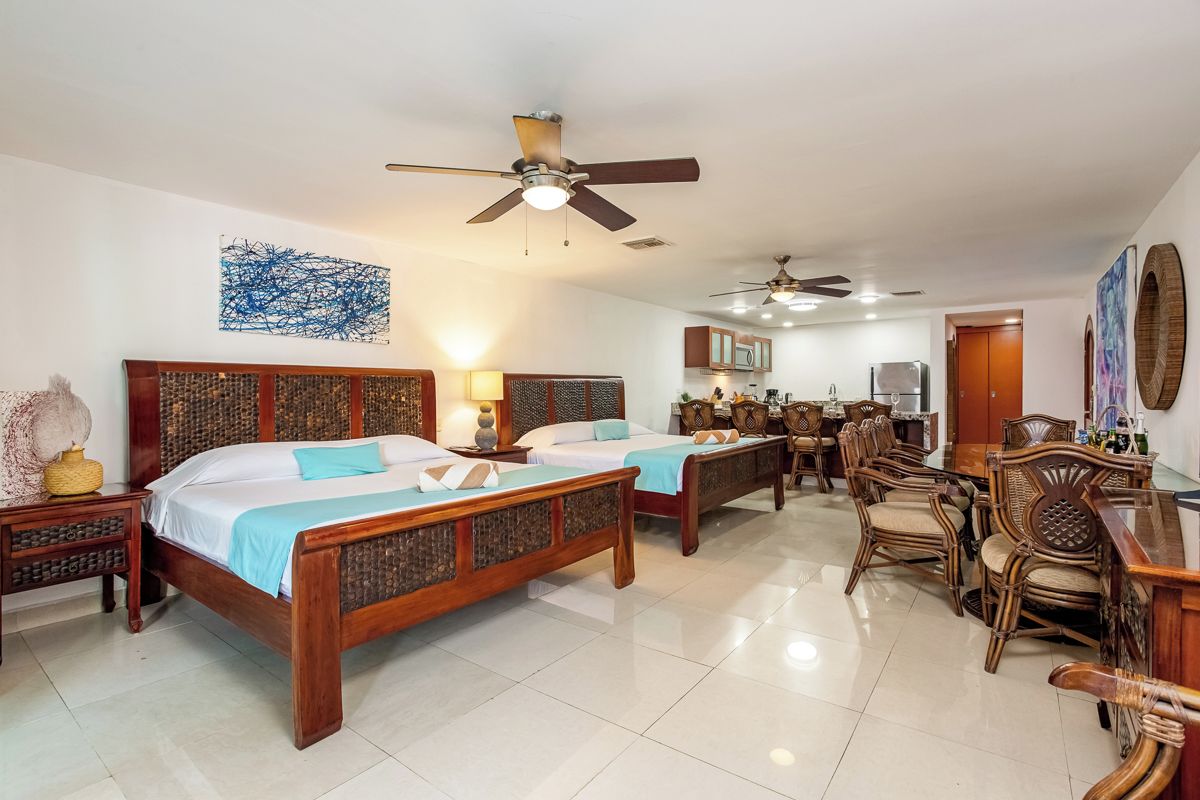 Quarto de hotel com duas camas de madeira, área de refeições com cadeiras de vime e cozinha ao fundo.