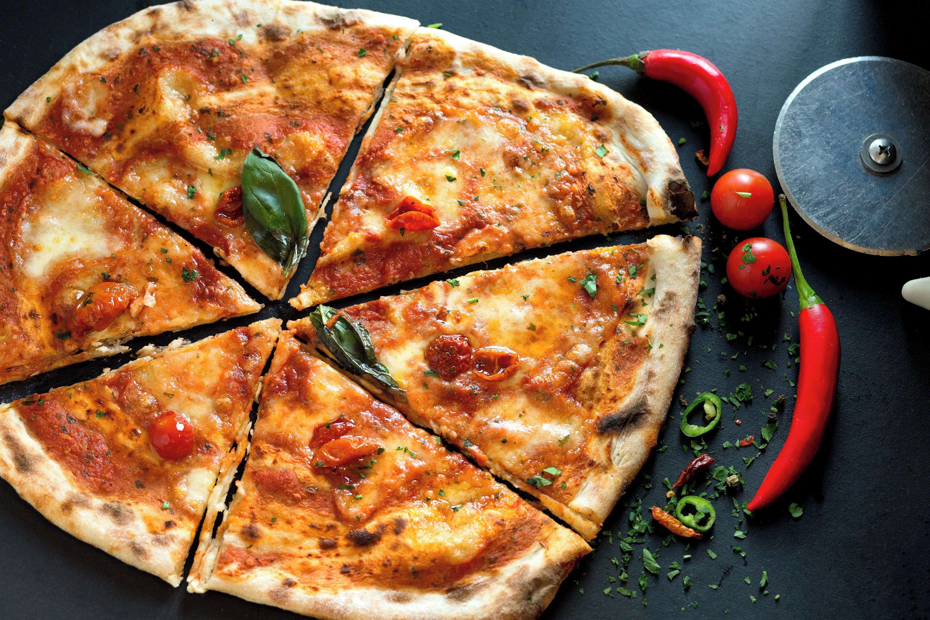 Pizza com oito fatias, decorada com ervas e tomates, rodeada por pimentas vermelhas e um cortador de pizza sobre uma superfície escura.