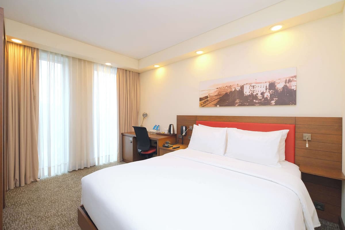 Quarto de hotel com uma cama grande, uma escrivaninha com cadeira, telefone e um quadro na parede.