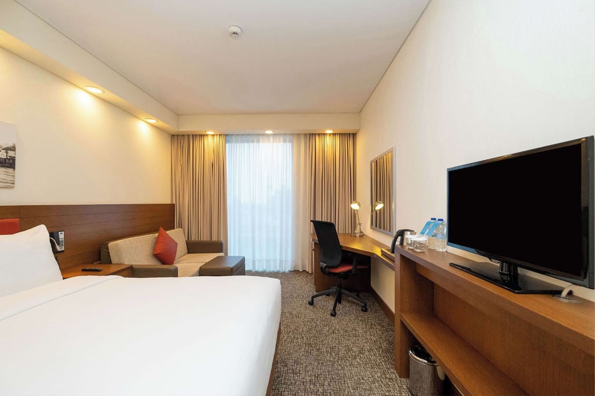 Quarto de hotel com cama, escrivaninha com computador, televisão e janela com cortinas.
