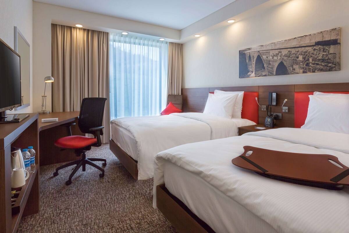 Quarto de hotel com duas camas, uma escrivaninha com cadeira, televisão e um quadro na parede.