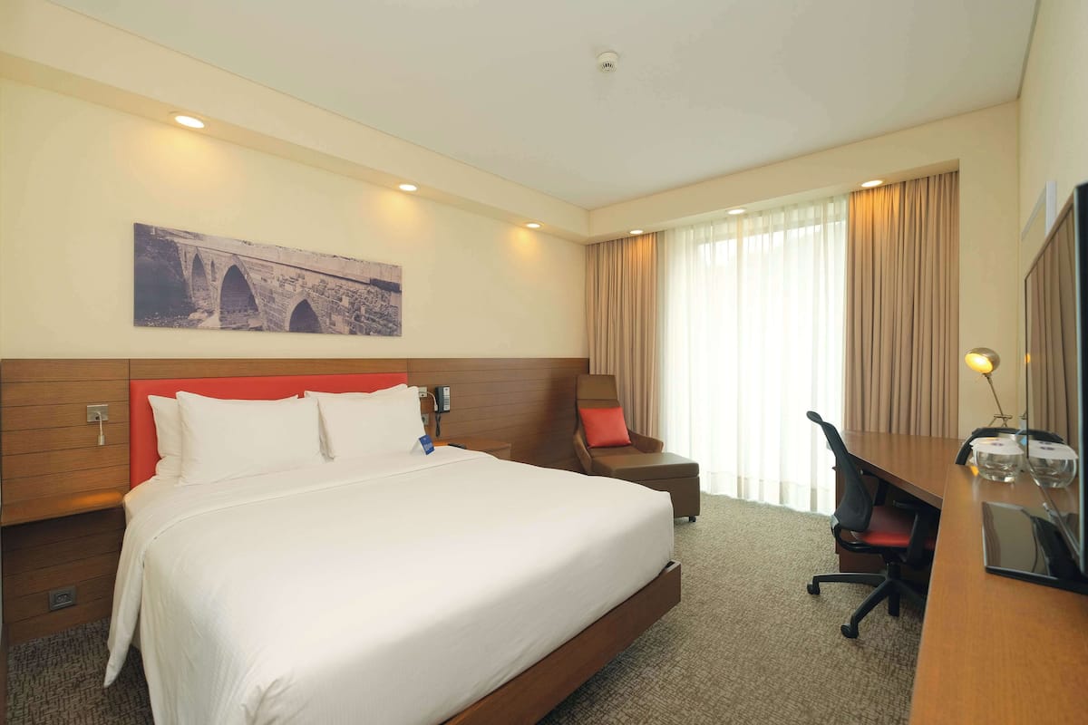 Quarto de hotel com uma cama grande, uma escrivaninha com cadeira, uma TV e um quadro na parede.