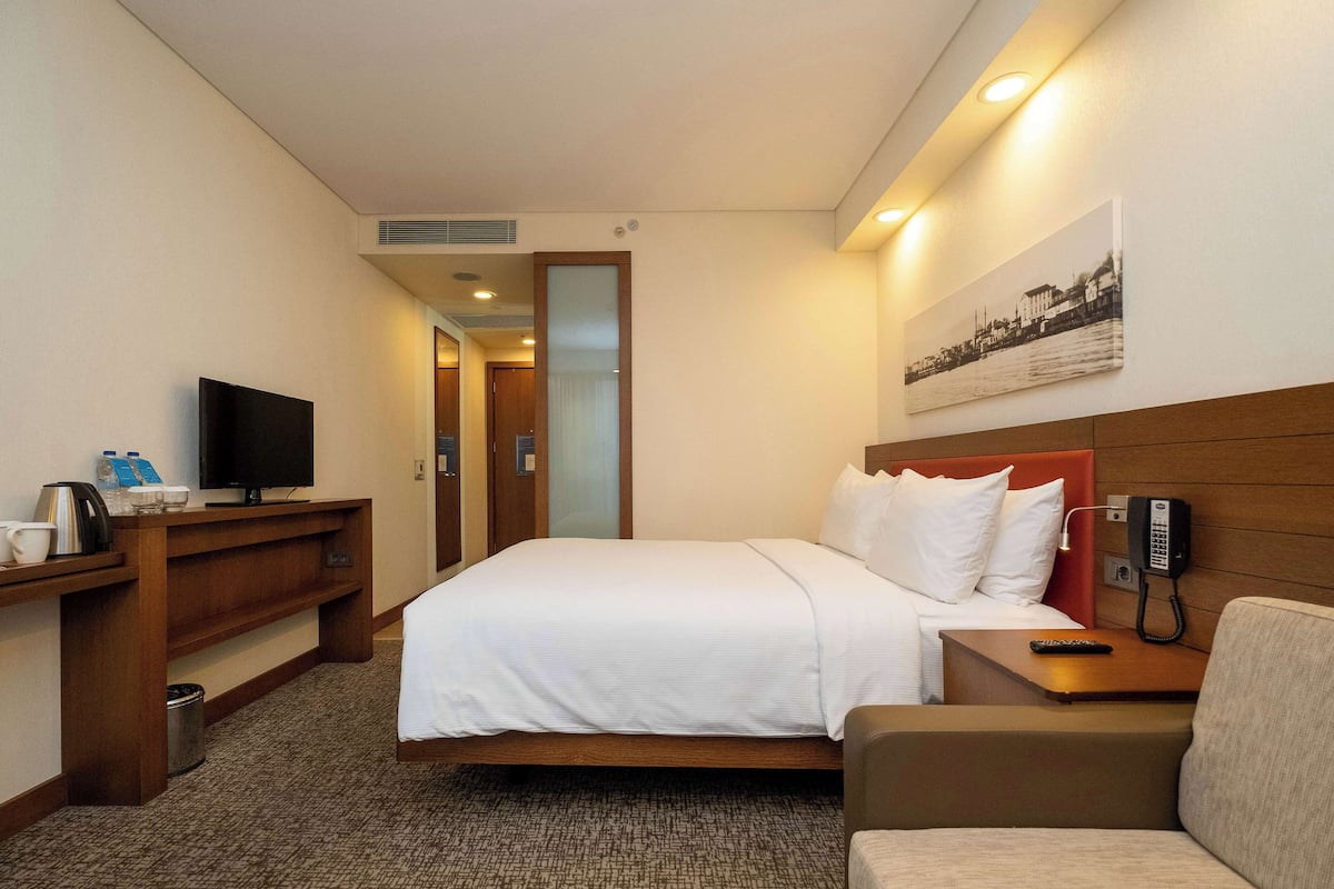 Quarto de hotel com uma cama, televisão, escrivaninha com cafeteira, telefone e um quadro na parede.