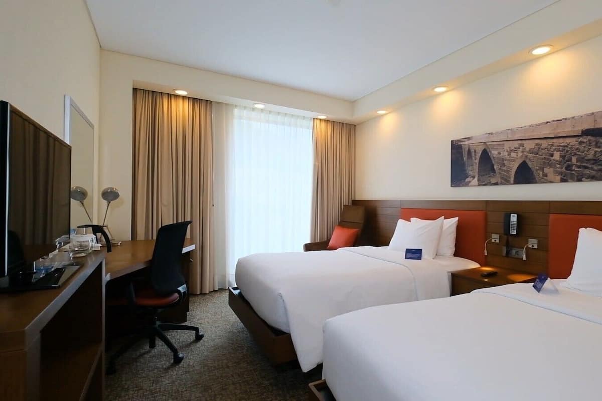 Quarto de hotel com duas camas, uma escrivaninha, uma cadeira, uma televisão e um quadro na parede.