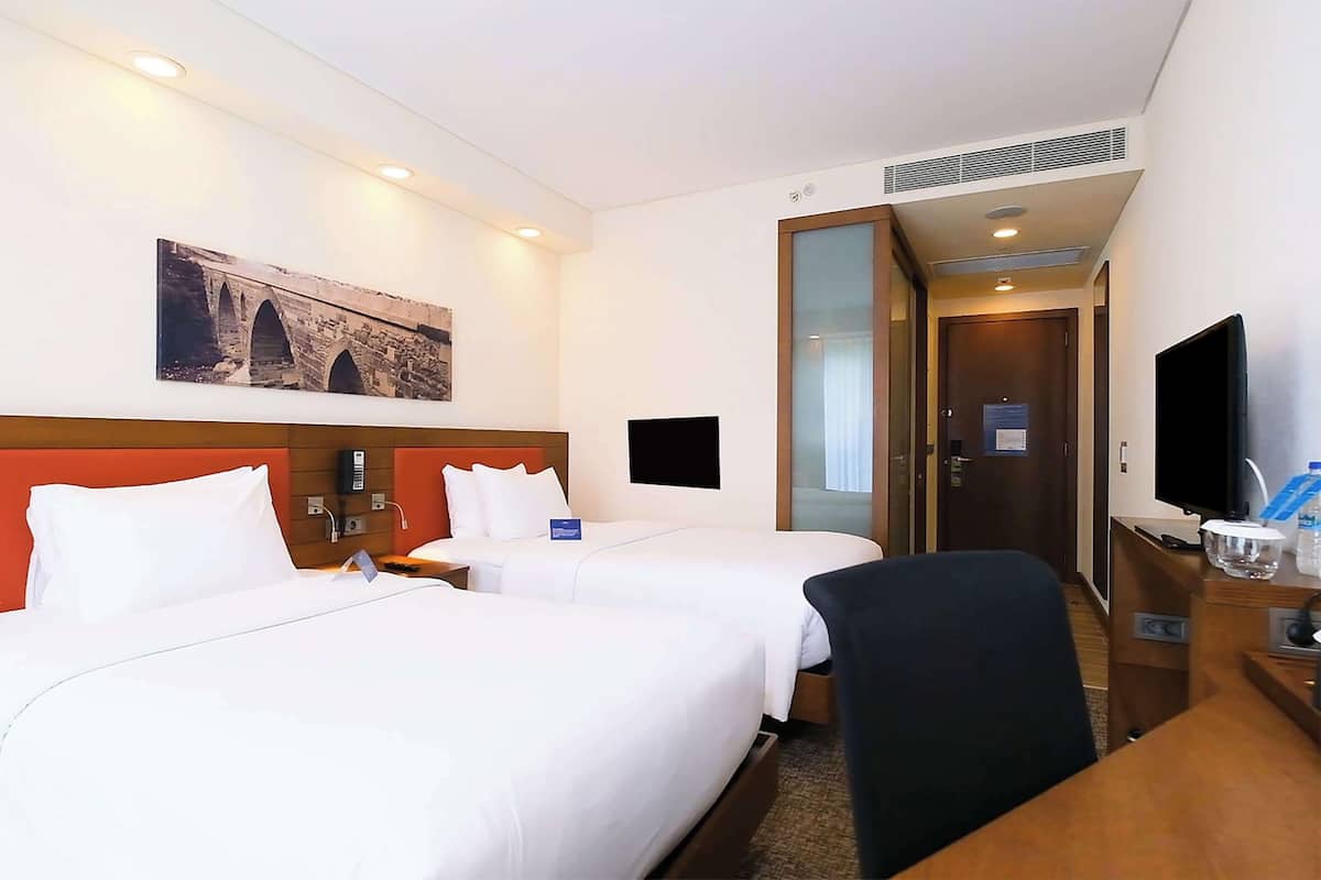 Quarto de hotel com duas camas, uma mesa com computador, televisão e um quadro na parede.