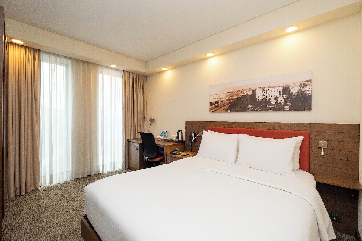 Quarto de hotel com uma cama grande, uma escrivaninha com cadeira, telefone e um quadro na parede.