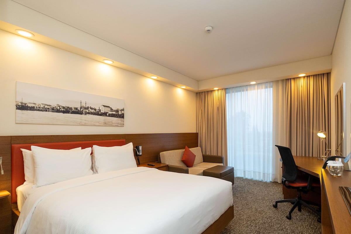 Quarto de hotel com uma cama grande, uma escrivaninha com cadeira, um sofá e uma janela ampla com cortinas.