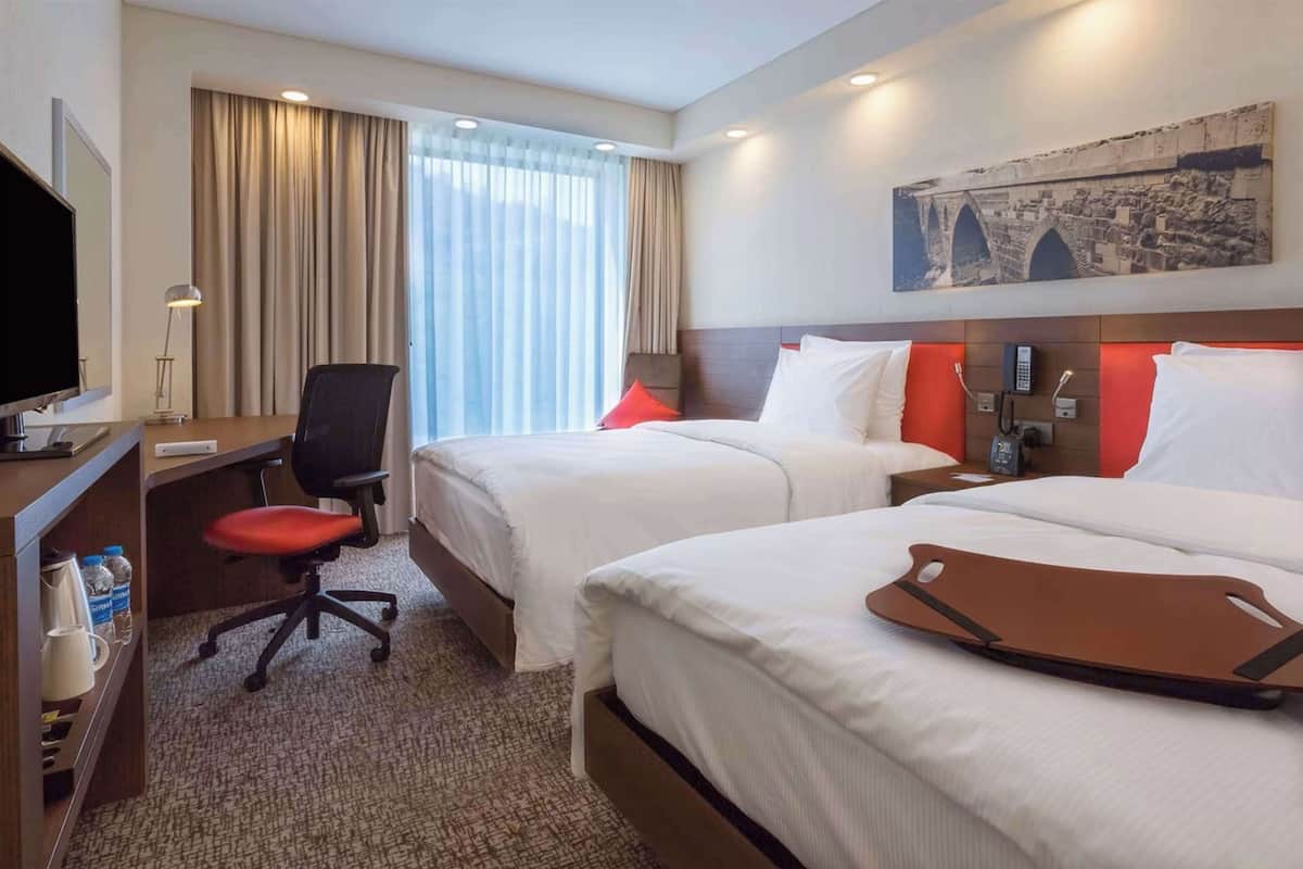Quarto de hotel com duas camas, uma escrivaninha com cadeira, televisão e um quadro na parede.