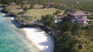 Vista aérea de uma propriedade costeira com praia, águas cristalinas e uma casa grande com piscina.