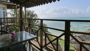 Um deck de madeira com mesa e cadeiras de vidro, com vista para uma praia e águas turquesa cristalinas e uma palmeira.