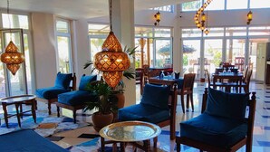 Um hall amplo com assentos estofados em azul, uma planta em vaso central, luminárias decorativas pendentes e vista para a área externa através de grandes janelas.