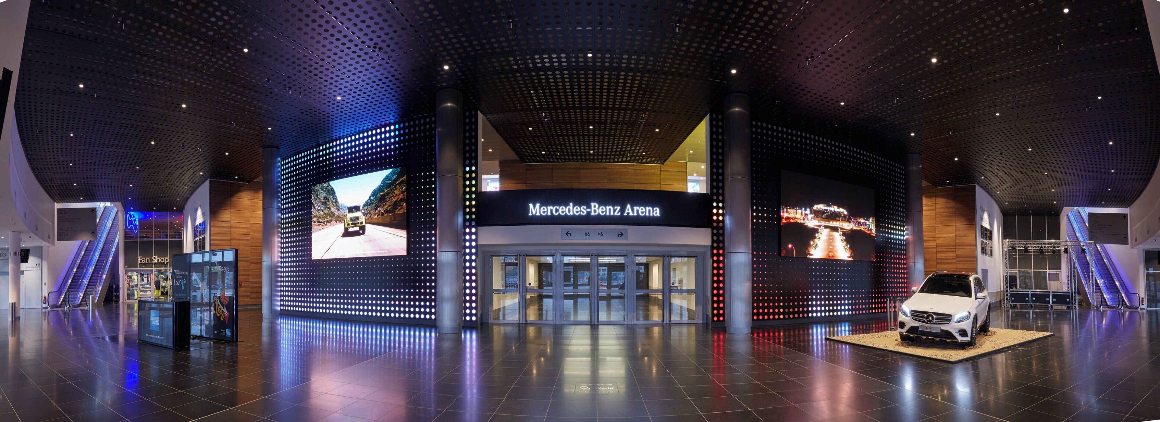 Espaço moderno em ambiente interno com a placa Mercedes-Benz Arena, escadas rolantes e a exposição de um carro branco.