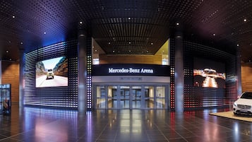 Espaço moderno em ambiente interno com a placa Mercedes-Benz Arena, escadas rolantes e a exposição de um carro branco.