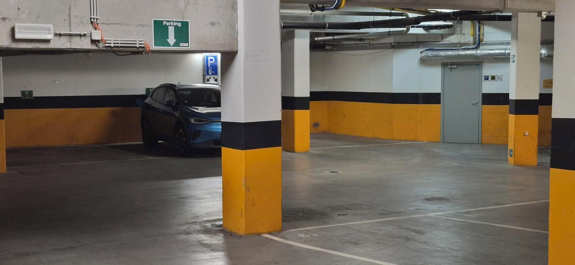 Garagem subterrânea de estacionamento com pilares amarelos e pretos, um carro azul estacionado e uma placa de estacionamento verde.