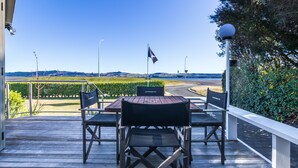 Um deck de madeira com mesa e cadeiras, com vista para uma paisagem panorâmica com uma bandeira, um poste de luz e uma sebe.