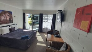 Quarto de hotel com cama, uma mesinha, uma cadeira, televisão e um quadro na parede.