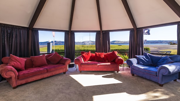 Quarto espaçoso com teto alto e grandes janelas que oferecem vista para uma paisagem verde e um corpo d'água. Há três sofás com almofadas vermelhas e azuis.