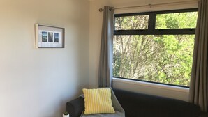 Um quarto aconchegante com um sofá cinza escuro, uma pequena mesa redonda com uma revista, um quadro na parede e uma janela com vista para árvores.