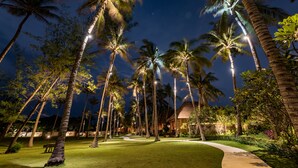 Vista noturna de um resort com palmeiras, um caminho e um edifício com telhado de palha.