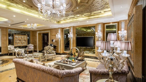 Uma sala de estar luxuosa com lustres elaborados, assentos confortáveis, uma grande televisão de tela plana e um piano de cauda.