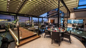 Uma área de refeições no terraço com mesas e cadeiras, piscina e vista para a paisagem urbana.
