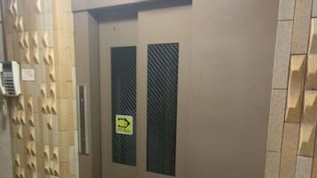 Um elevador com porta metálica, placa de sinalização e um cesto de lixo.