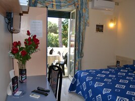 Quarto de hotel com uma cama, uma mesa com um vaso de rosas vermelhas, uma cadeira e vista para o exterior através de uma porta de vidro.