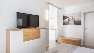 Quarto moderno com televisão sobre um armário de madeira, cama com manta da marca, mesa de cabeceira de madeira pequena e pintura de um cais na parede.