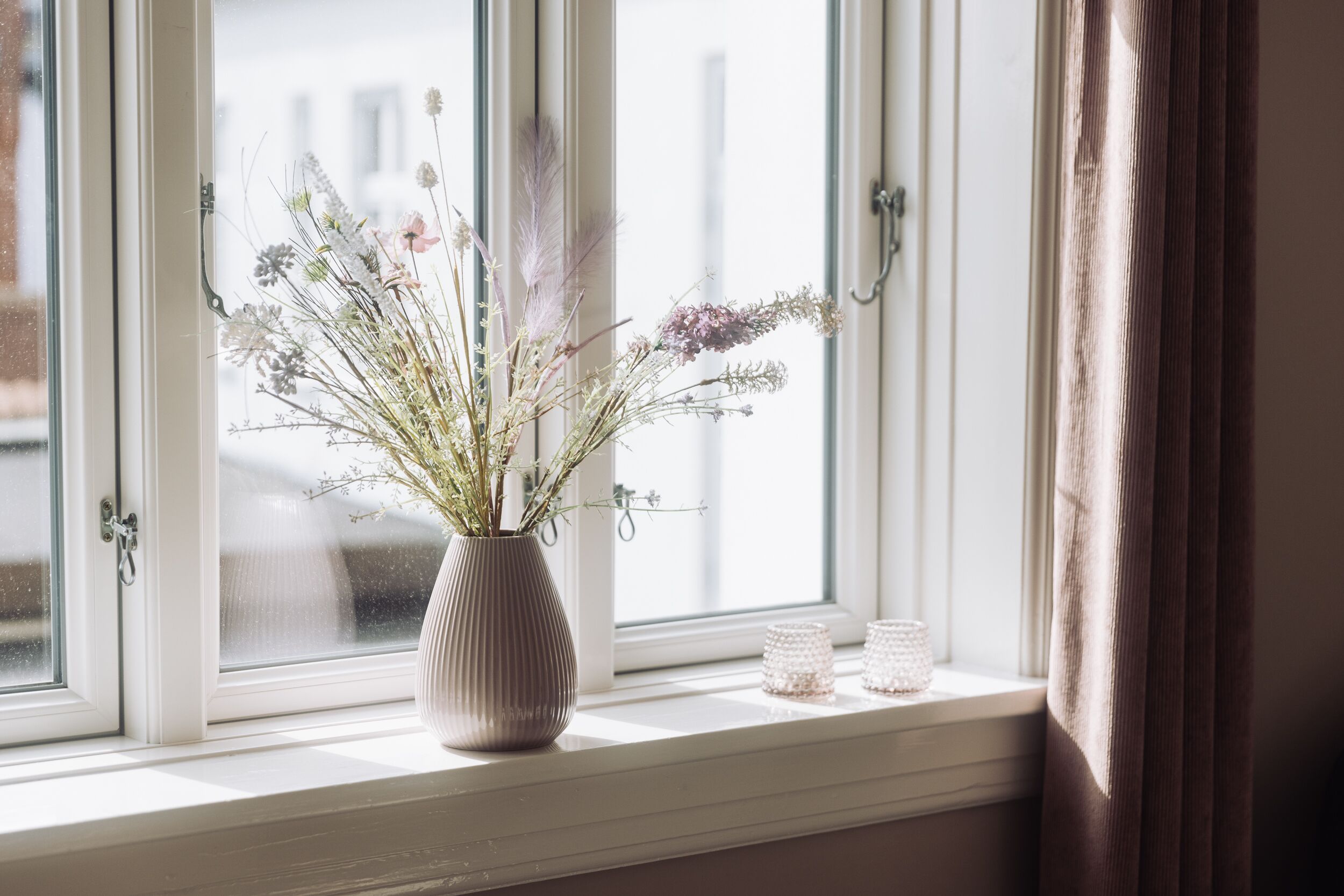 Uma janela com um vaso de flores secas e dois castiçais em um parapeito branco.