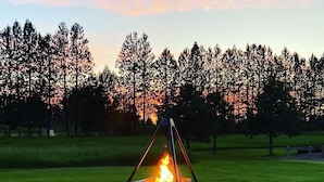 Montagem de fogueira com suporte de metal, rodeada por pedras e toras, com um campo gramado e árvores ao fundo durante o pôr do sol.