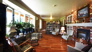 Uma sala de estar aconchegante com piso de madeira, lareira, estante de livros e uma janela grande com plantas.