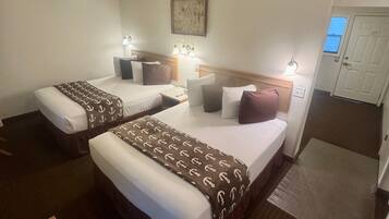 Quarto de hotel com duas camas, uma mesinha, uma cadeira e porta para um corredor.