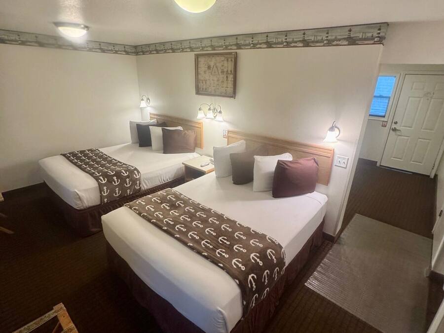 Quarto de hotel com duas camas, uma mesinha, uma cadeira e porta para um corredor.